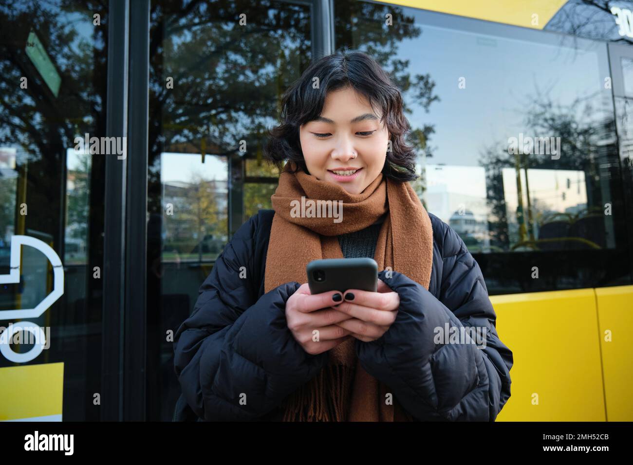 Beautiful korean girl, student on bus stop, looking at her smartphone ...