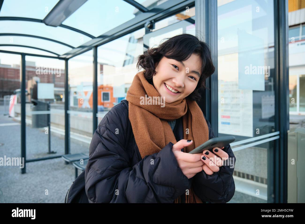 Cute smiling asian girl standing on bus stop, holding smartphone ...