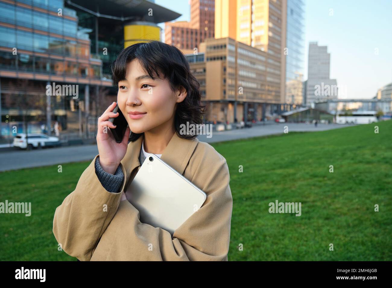 Stylish smiling girl, university student in trench coat, holds tablet ...