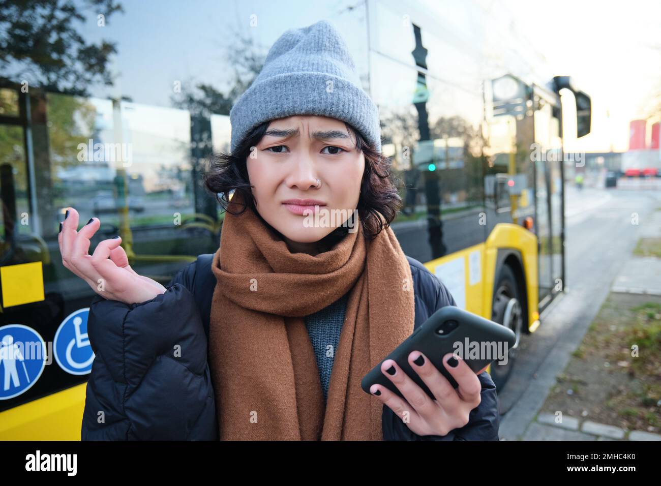 Portrait of confused asian girl, standing on bus stop, holding mobile ...