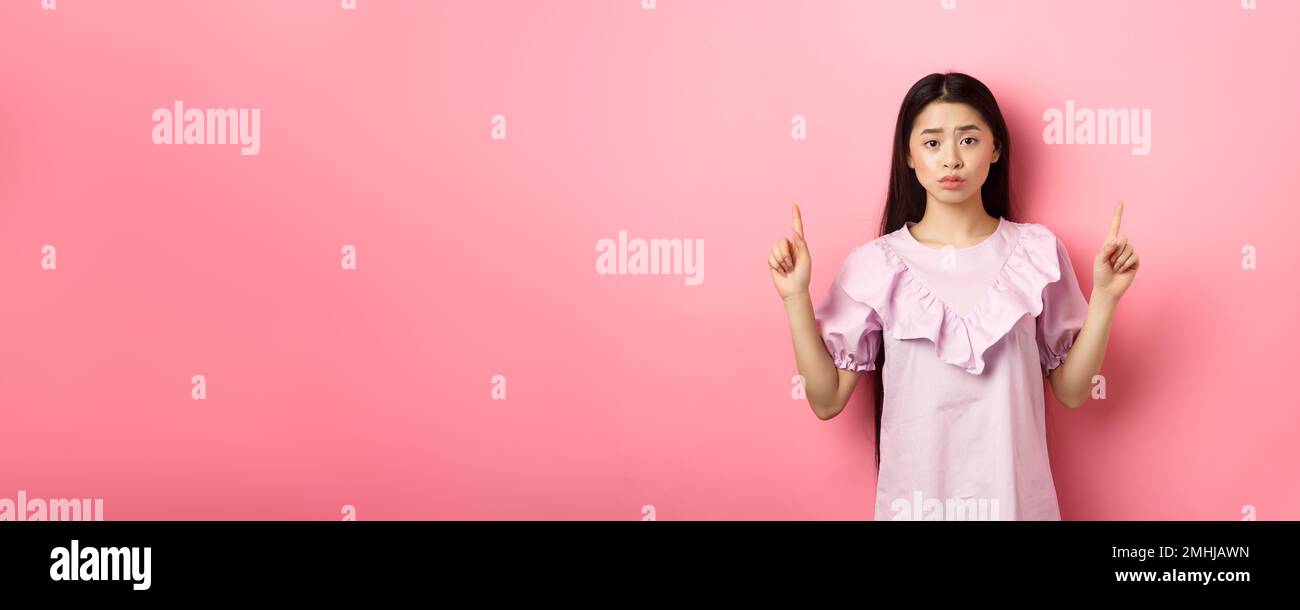 Cute timid asian woman pointing fingers up, frowning and look upset ...