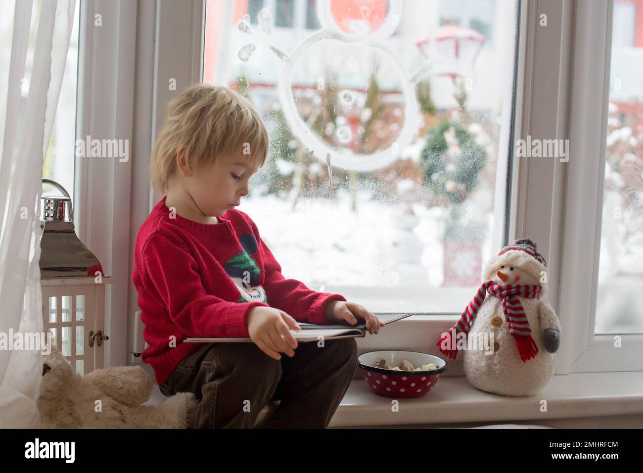 Cute blond toddler child, sitting on the window, looking at the snow ...