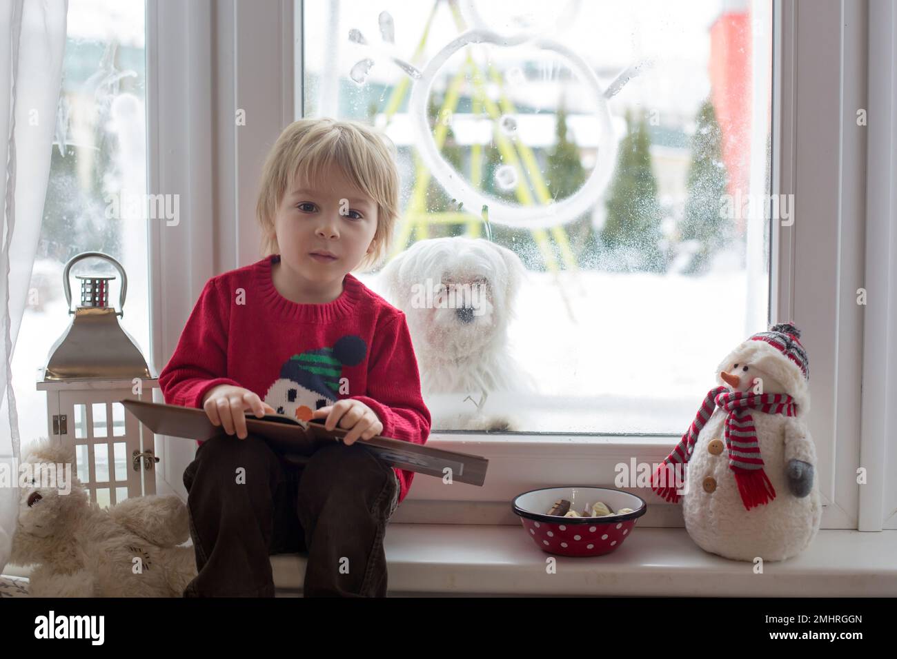 Cute blond toddler child, sitting on the window, looking at the snow ...