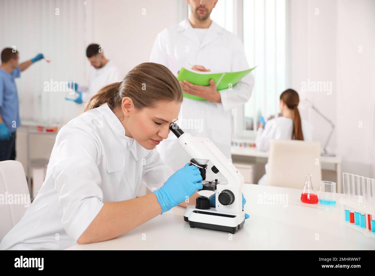 Scientist using microscope at table and colleagues in laboratory ...