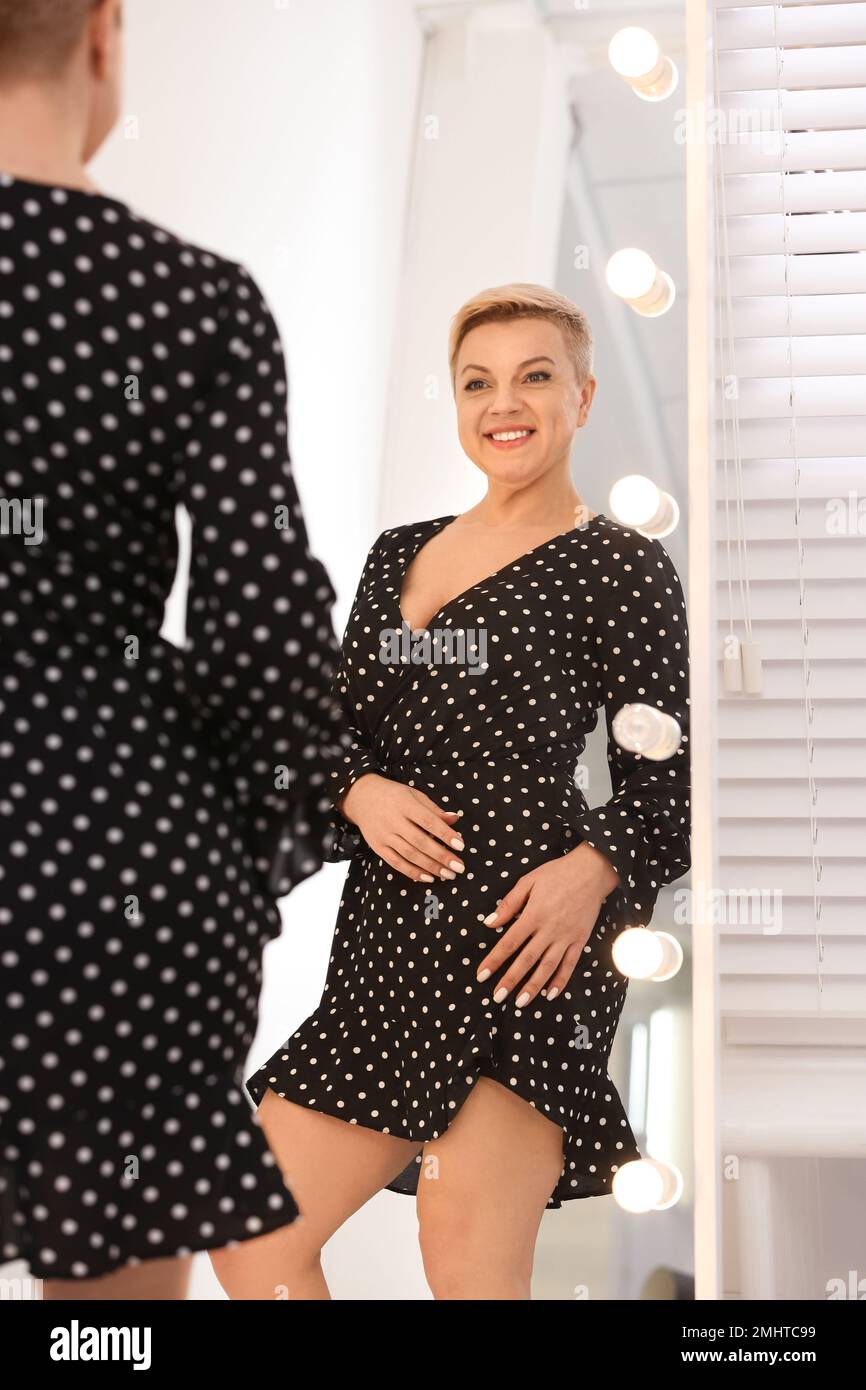 Beautiful mature woman looking at herself in large mirror with lights ...