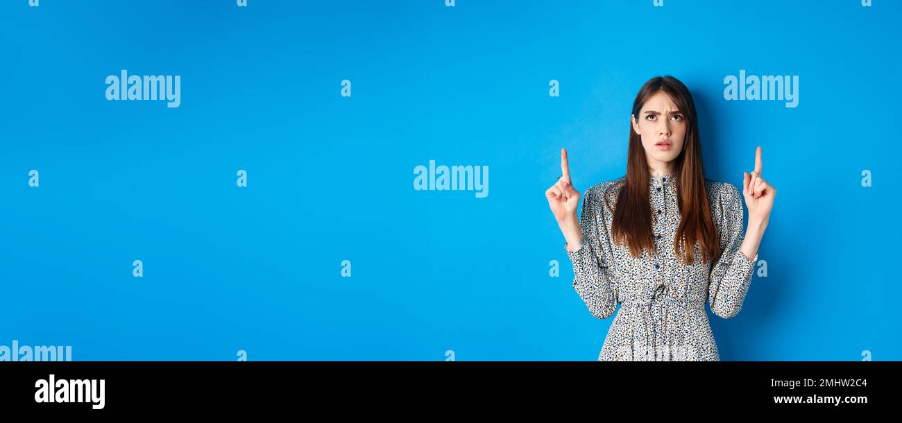 Confused attractive woman in dress frowning, pointing fingers up and ...