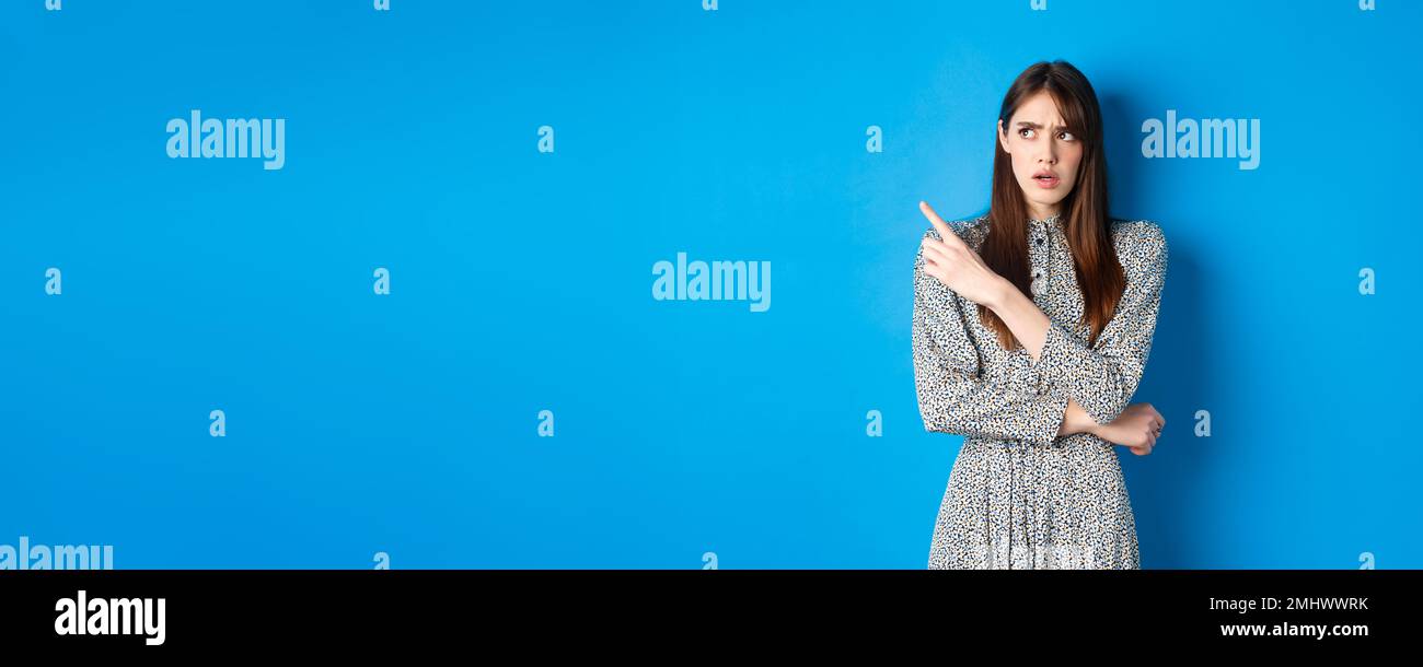 Confused girl in dress pointing and looking at upper left corner with ...