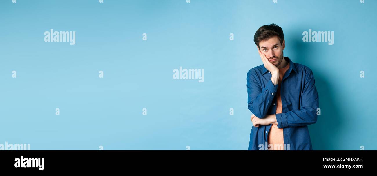 Sad and bored man lean face on hand, looking at camera empty-headed, standing bothered on blue ...