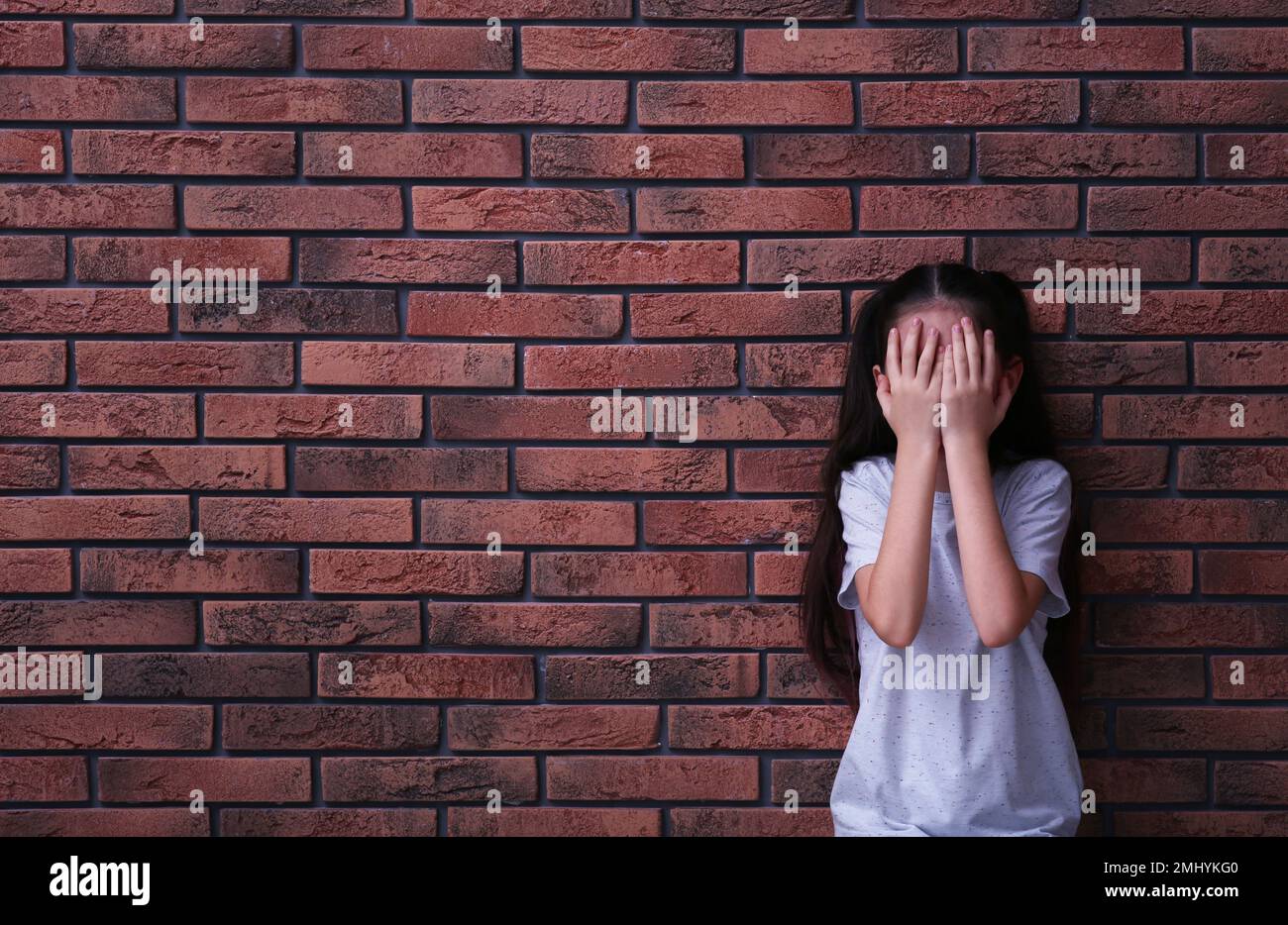Sad little girl closing eyes with hands near brick wall, space for text ...