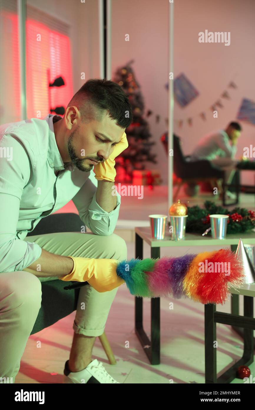 Man with dusting brush suffering from hangover in messy room after New ...