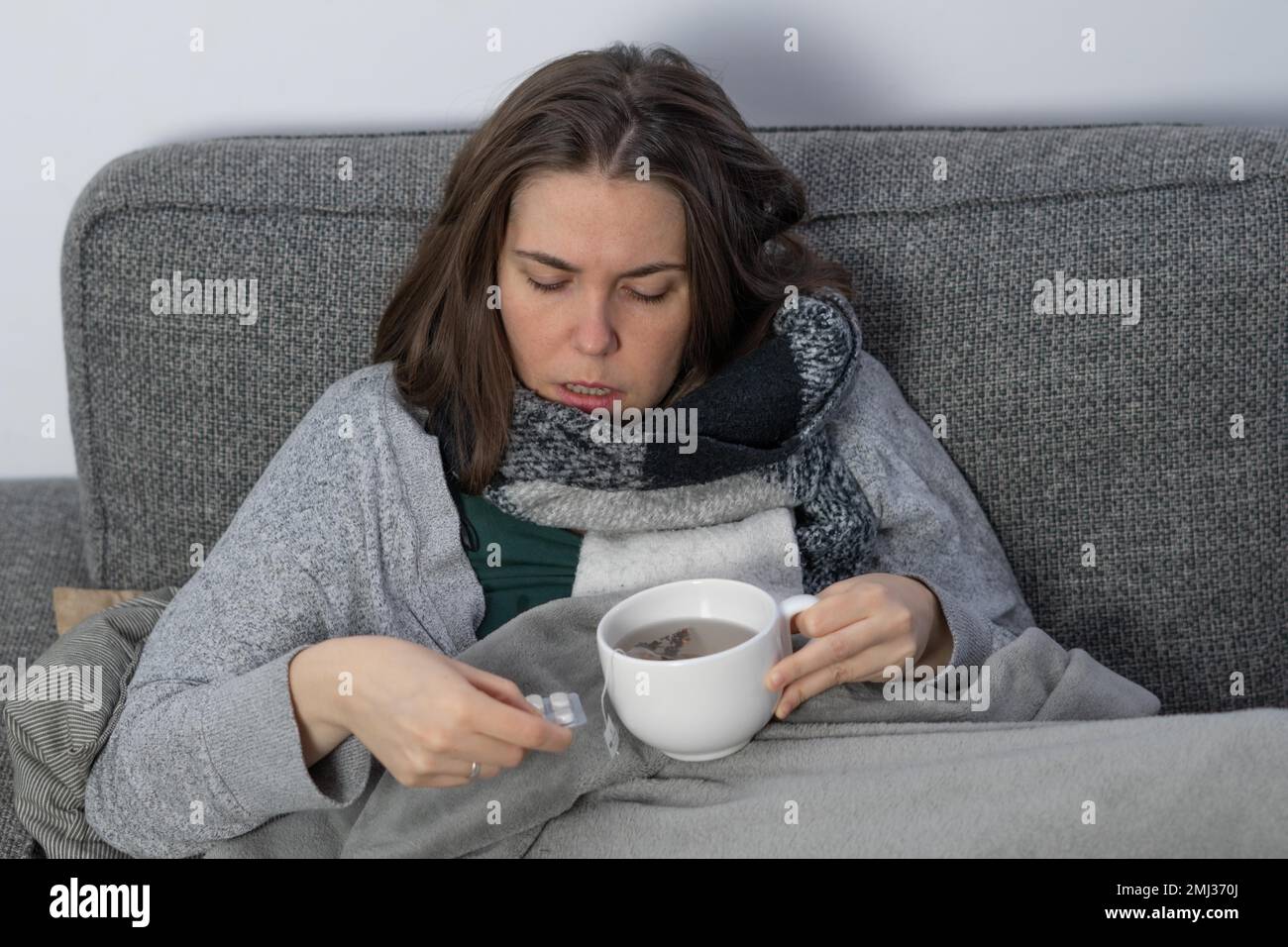 Sick woman covered with a blanket drinking an infusion on the couch in ...