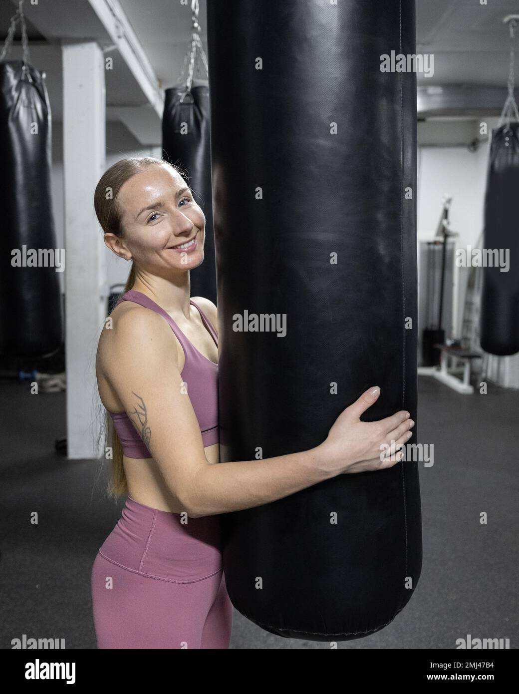 Long-haired blonde feminine girl boxer hugs a punching bag in a gym ...