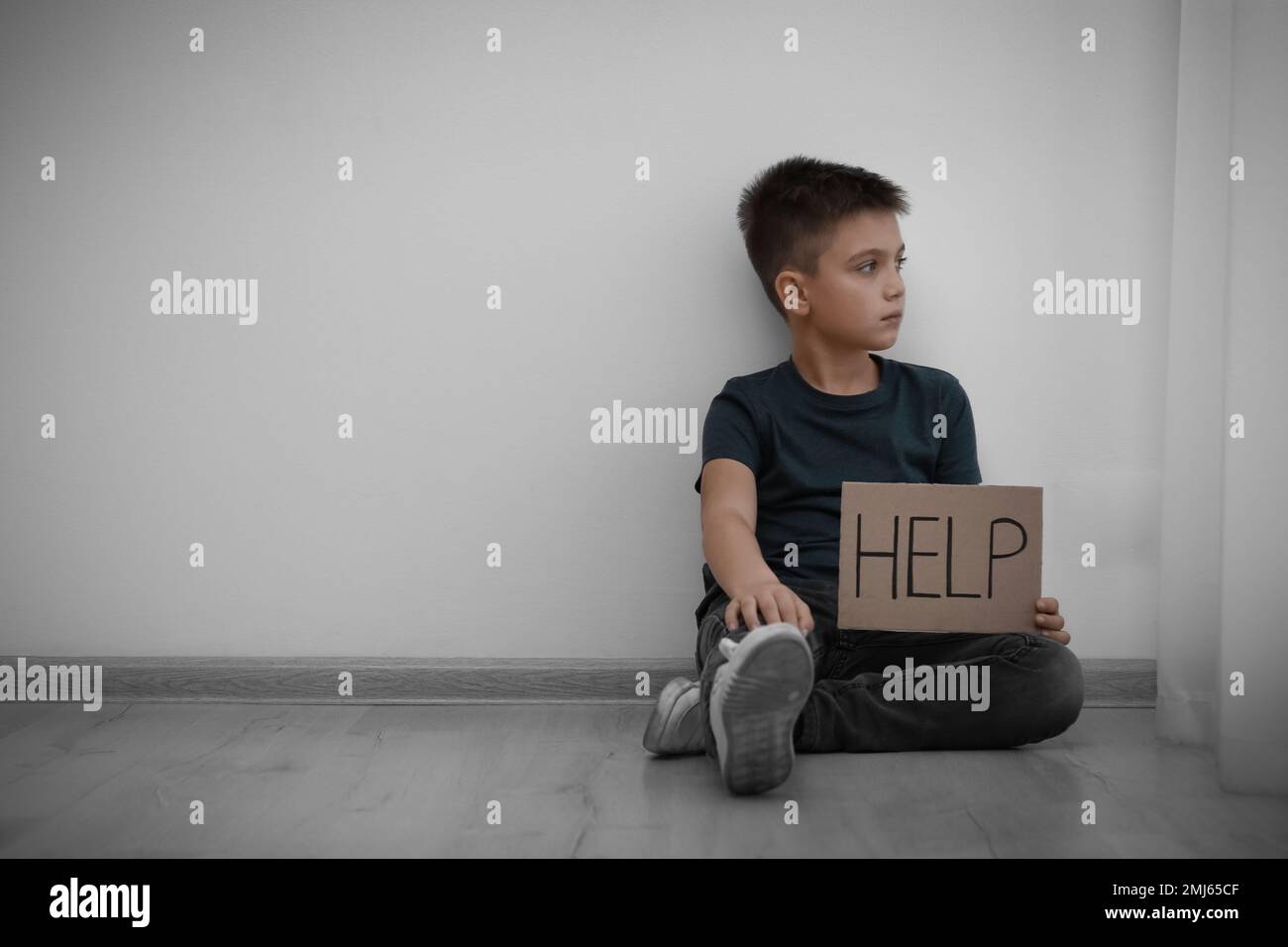 Sad little boy with sign HELP sitting on floor indoors, space for text ...