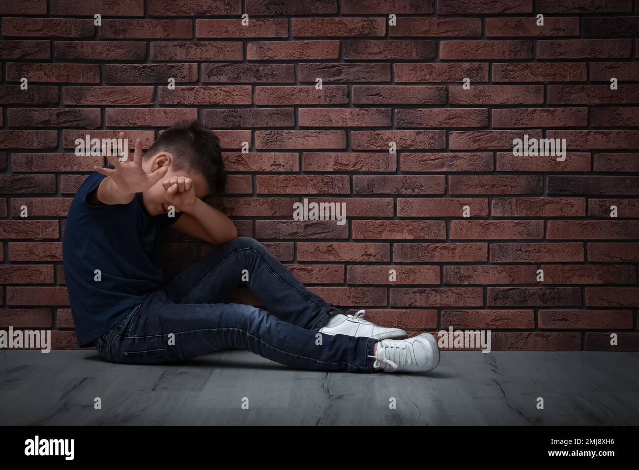 Scared little boy closing eyes with hand on floor near brick wall ...