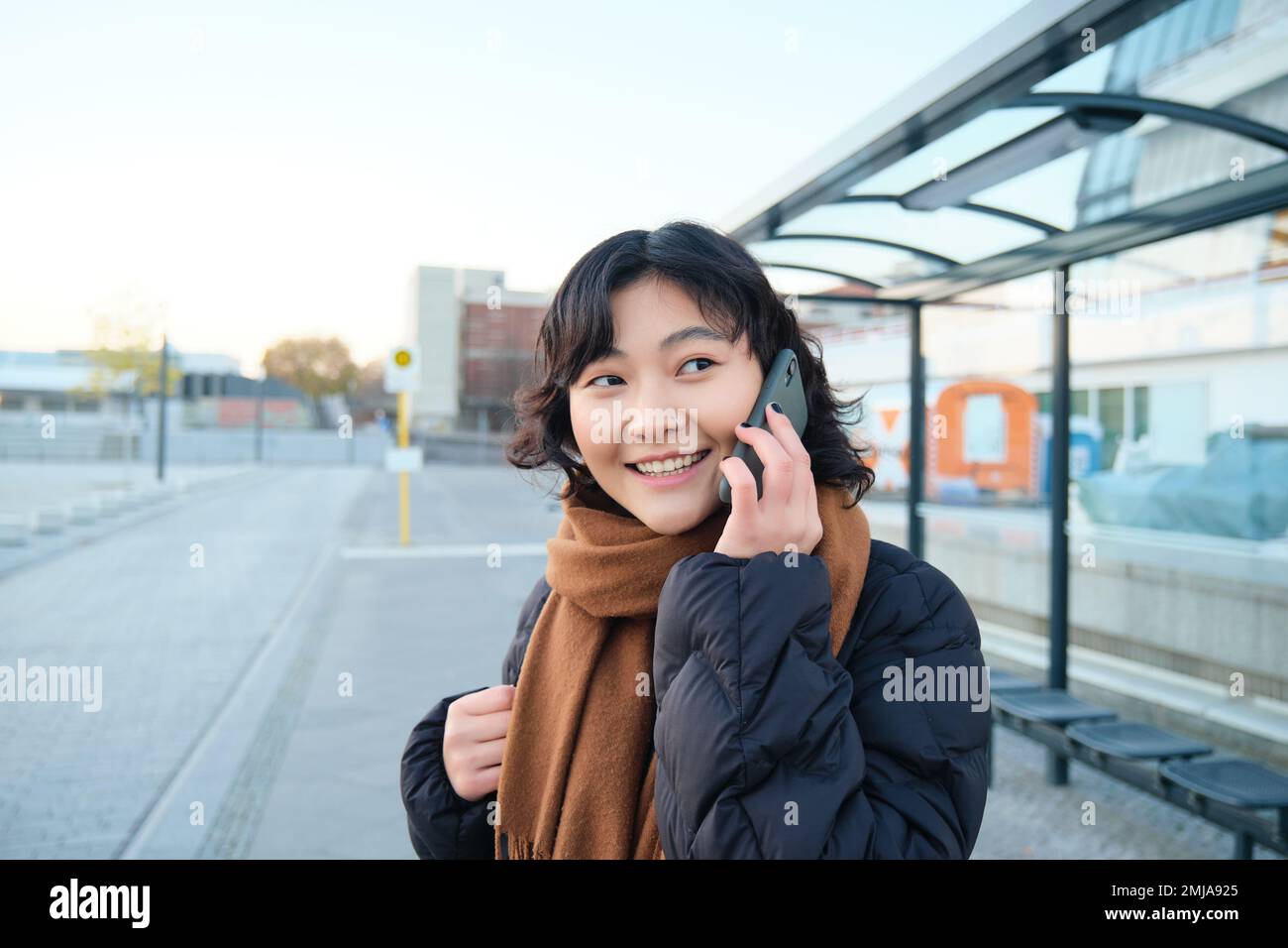 Beautiful smiling korean girl, waiting on bus stop, using public transport, talking on mobile ...