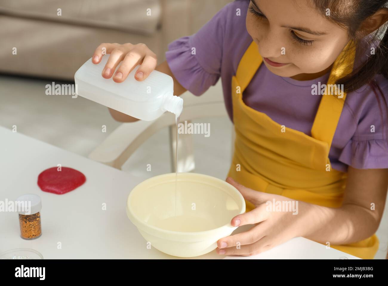 Cute little girl pouring glue into bowl at table in room. DIY slime toy ...