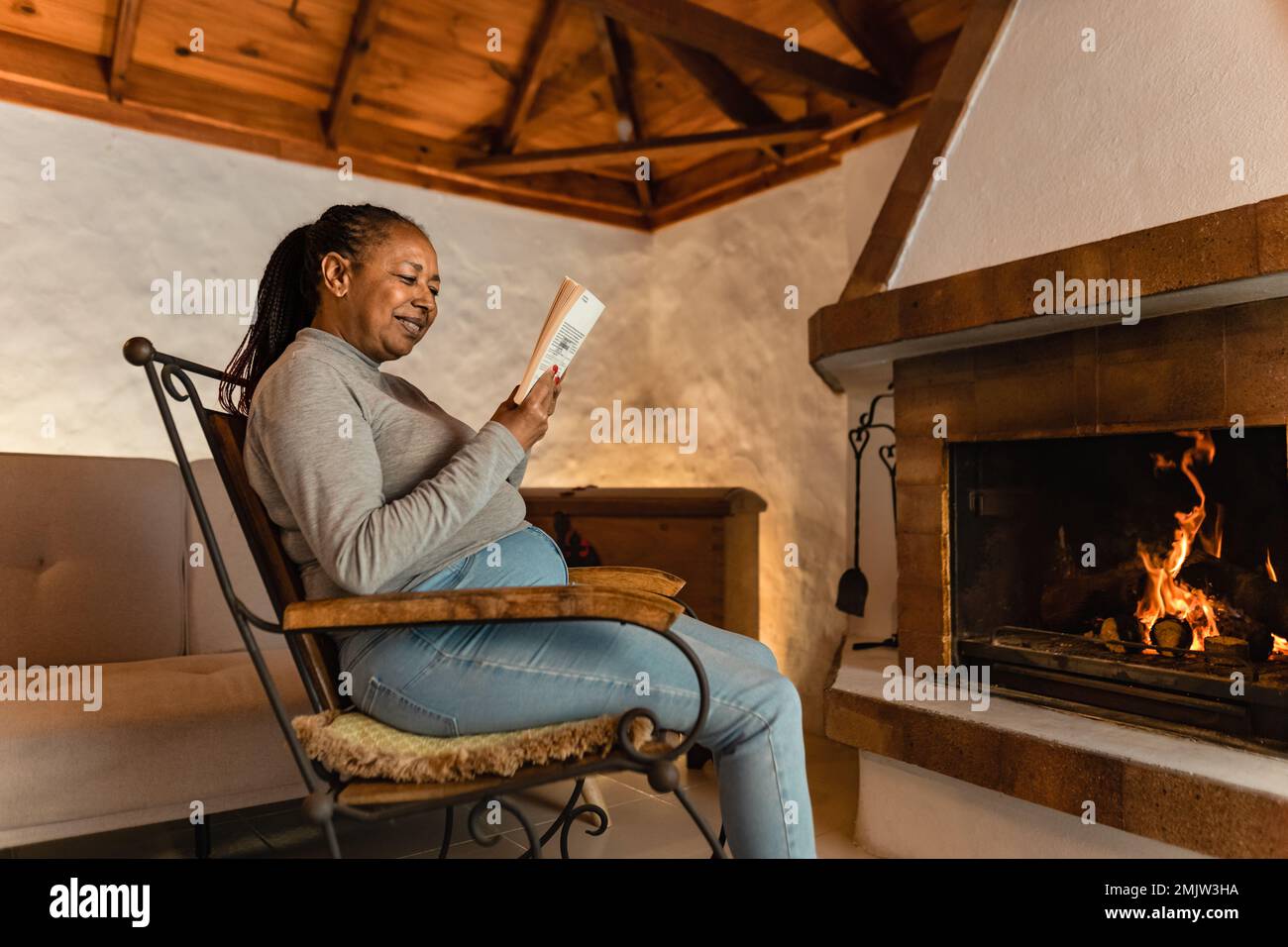 Senior African woman reading book while warming in front of fireplace ...