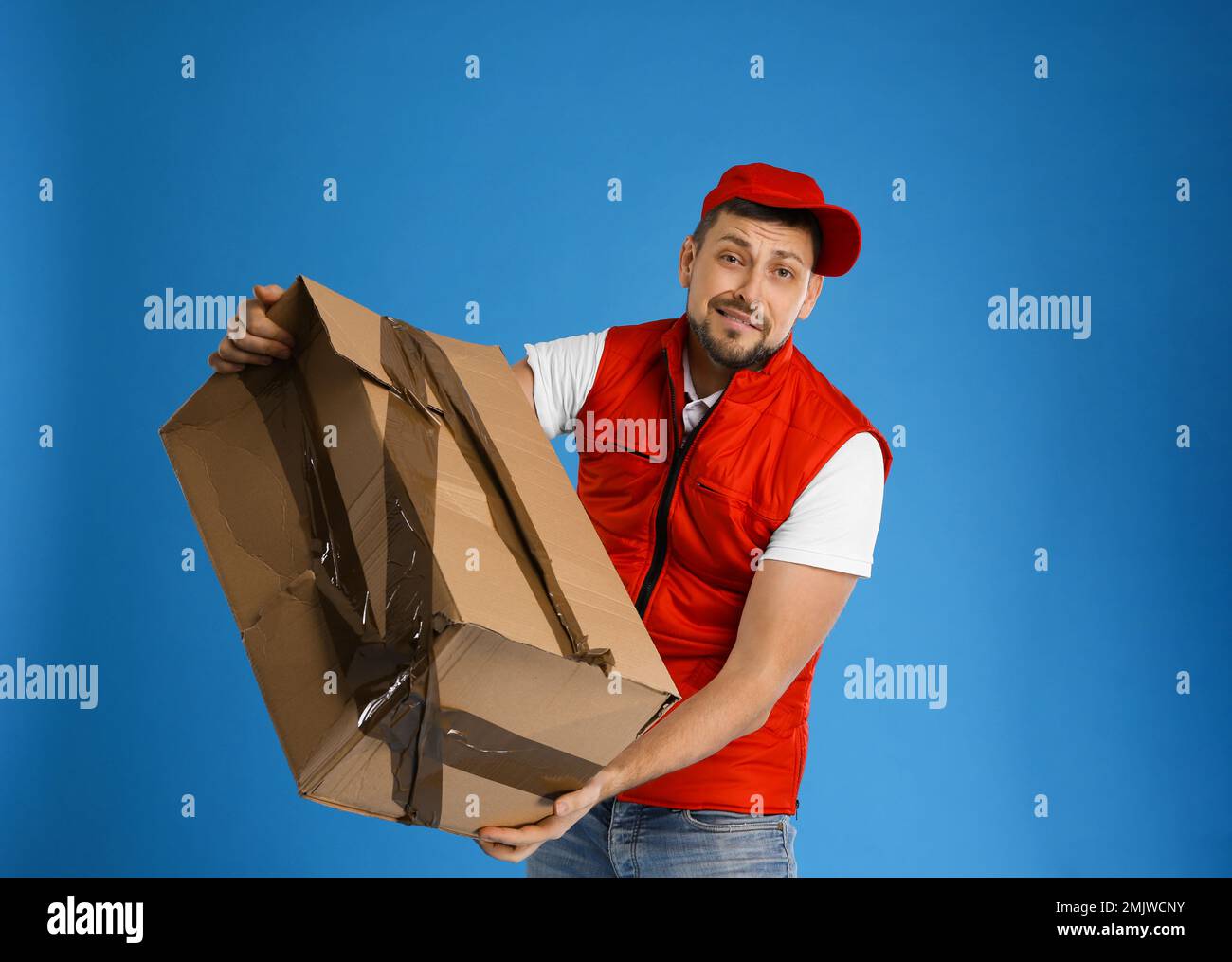 Emotional courier with damaged cardboard box on blue background. Poor ...