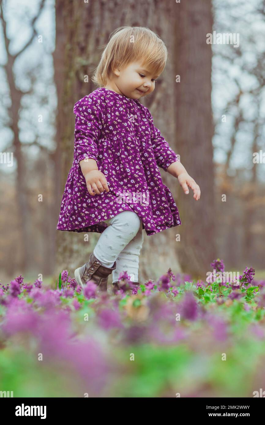 blond baby in a colored dress running in a forest glade with flowers ...