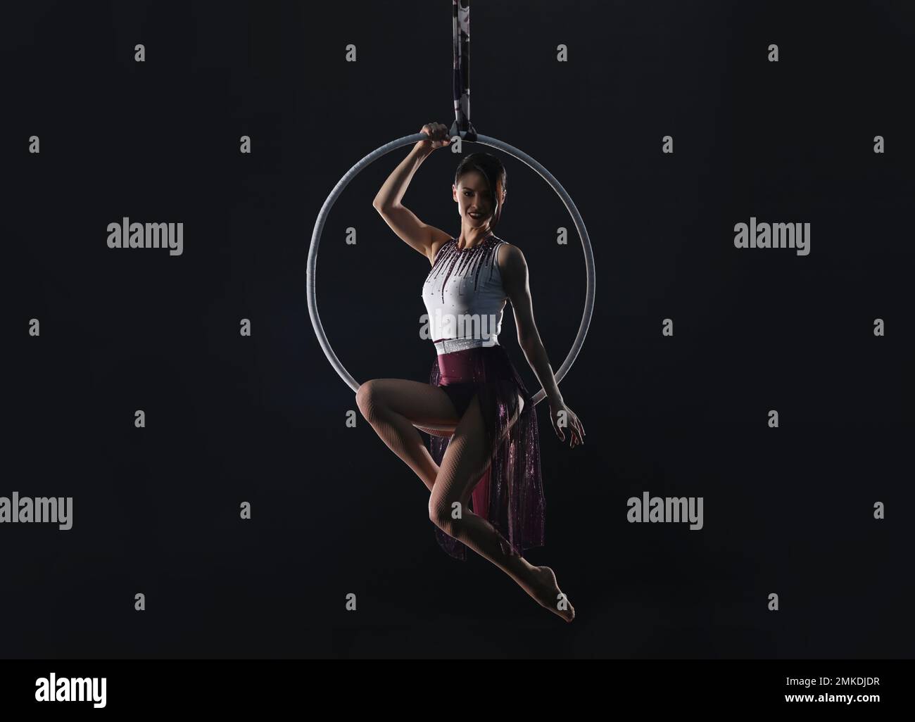 Young woman performing acrobatic element on aerial ring against dark ...