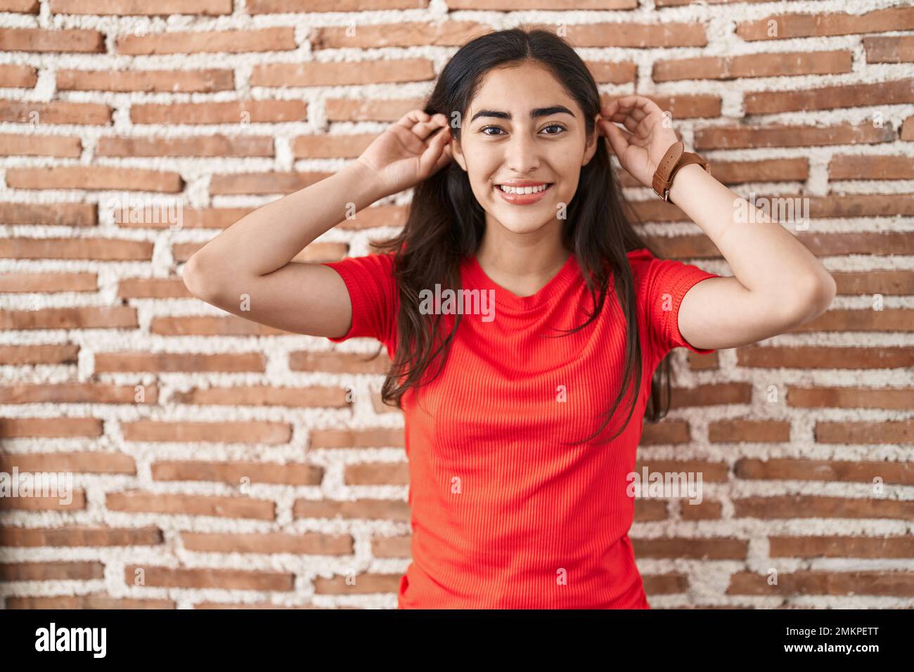 Young teenager girl standing over bricks wall smiling pulling ears with ...