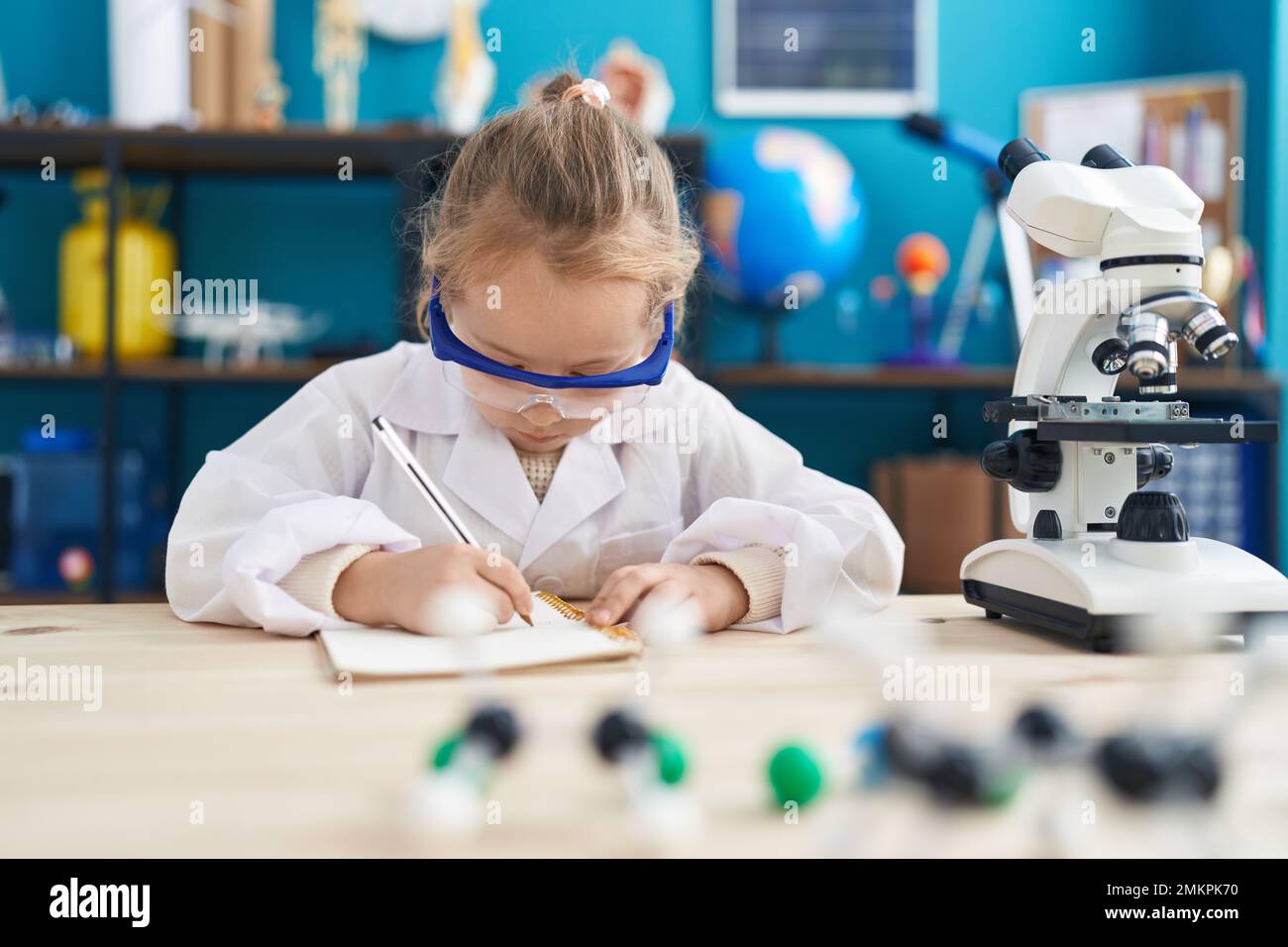 Adorable blonde girl student using microscope writing on notebook at ...