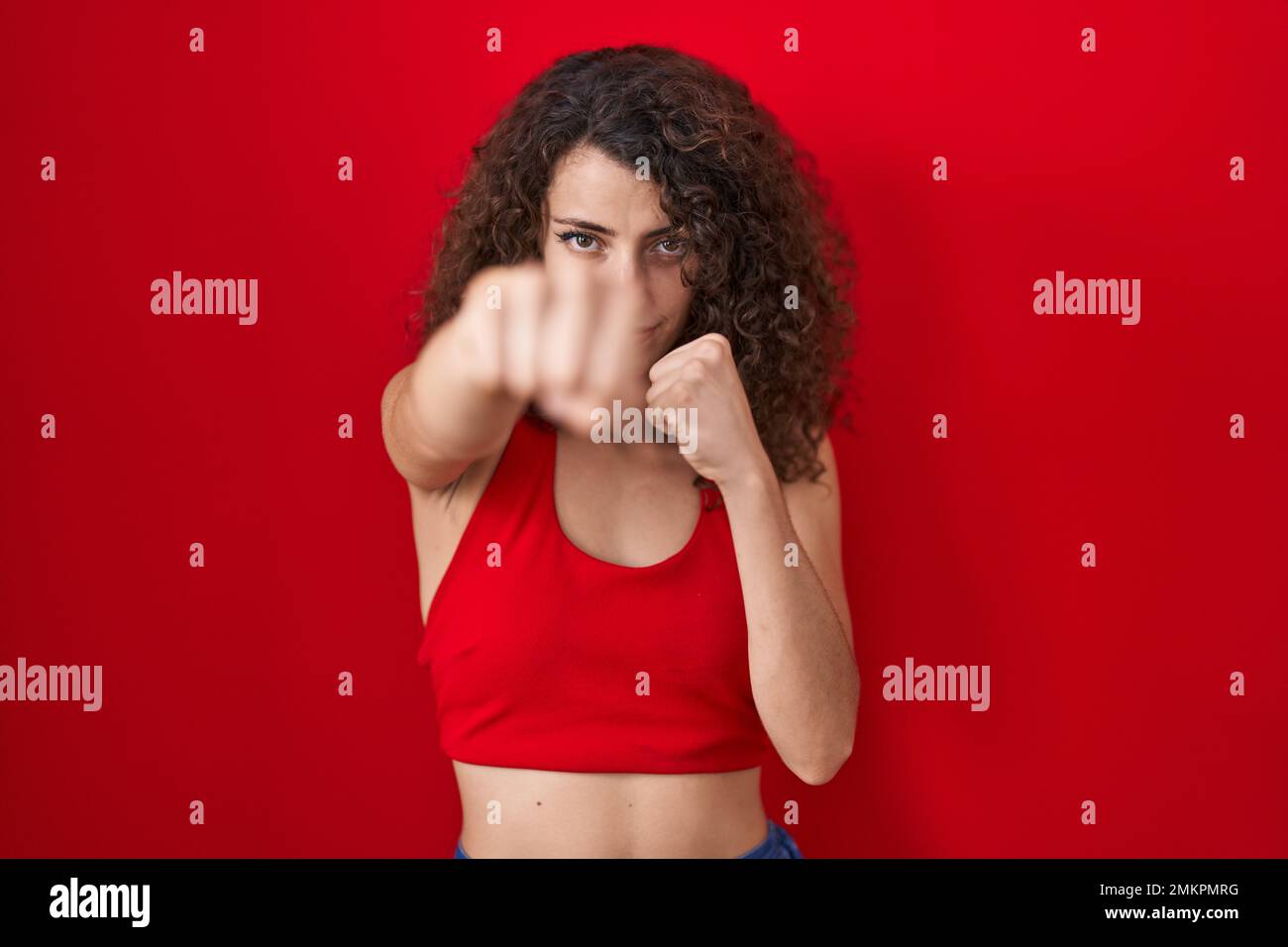Hispanic woman with curly hair standing over red background punching ...