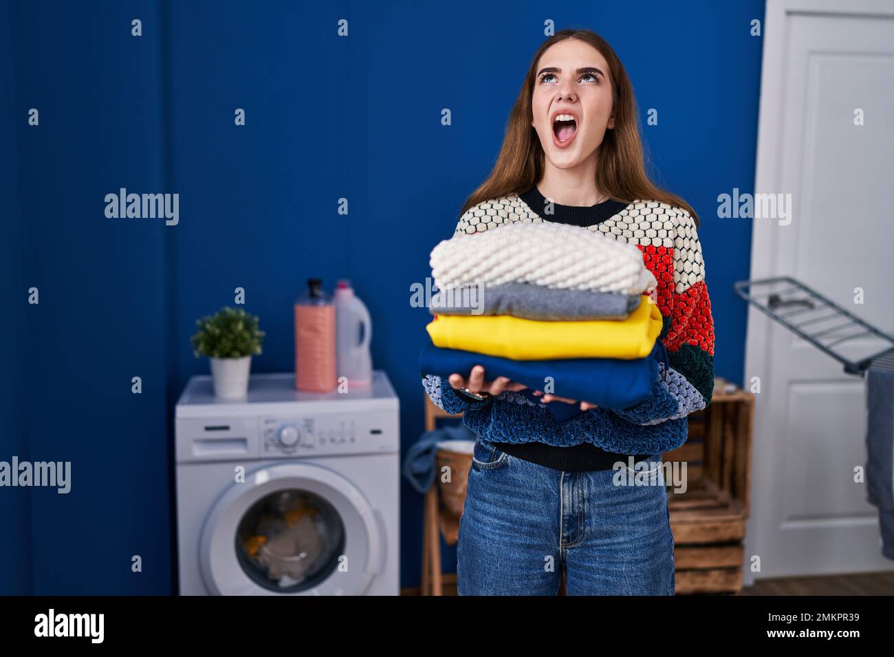Young hispanic girl holding clean laundry angry and mad screaming frustrated and furious ...