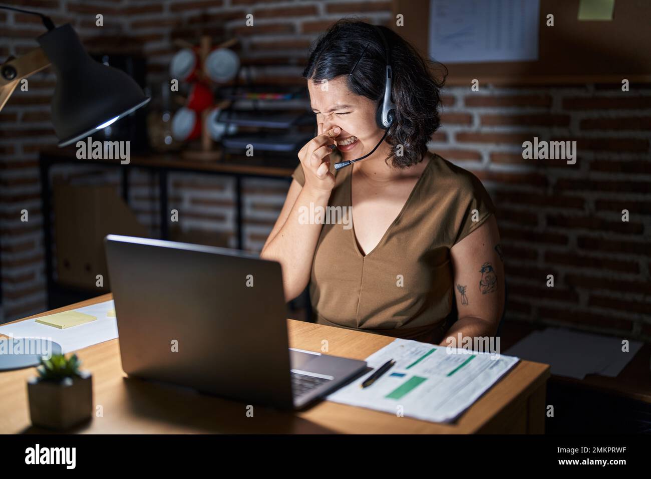 Young hispanic woman working at the office at night smelling something ...