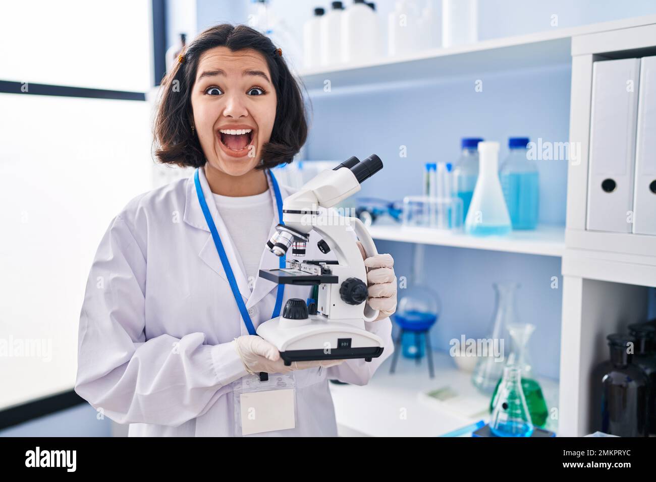 Young hispanic woman working at scientist laboratory holding microscope ...