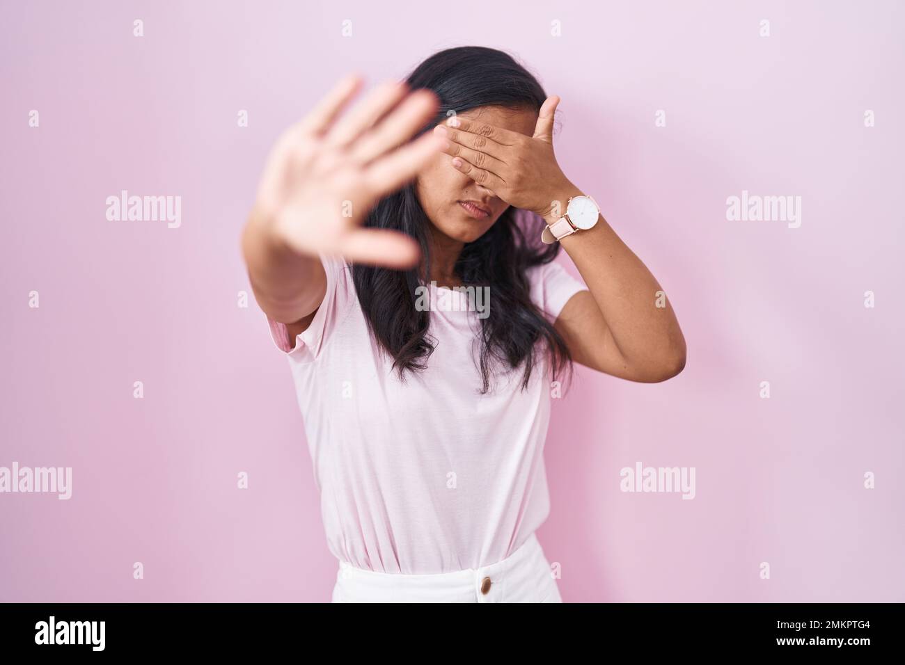 Young hispanic woman standing over pink background covering eyes with ...