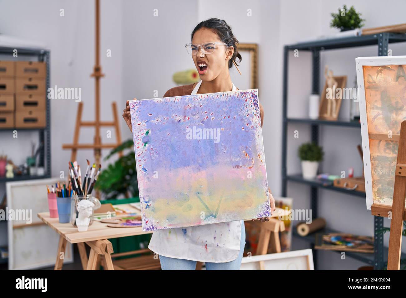 Young brazilian woman holding painter canvas at art studio angry and ...