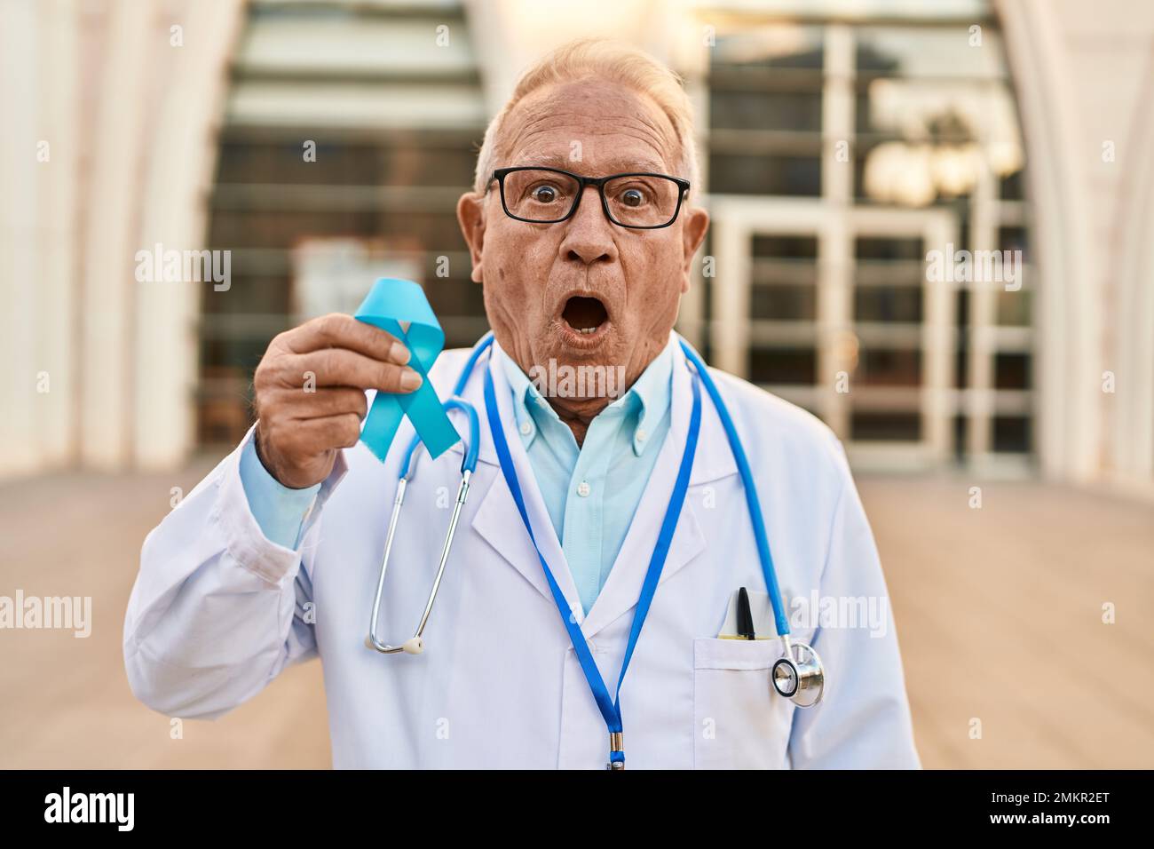 Senior doctor with grey hair holding blue ribbon scared and amazed with ...