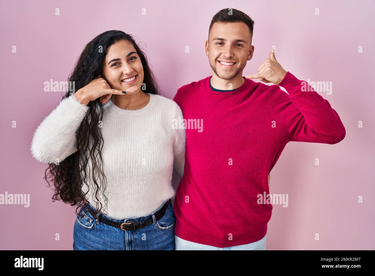 Young hispanic couple standing over pink background smiling doing phone ...