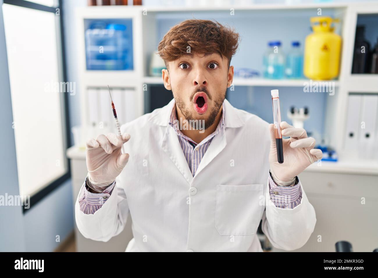 Arab man with beard working at scientist laboratory holding blood sample afraid and shocked with ...