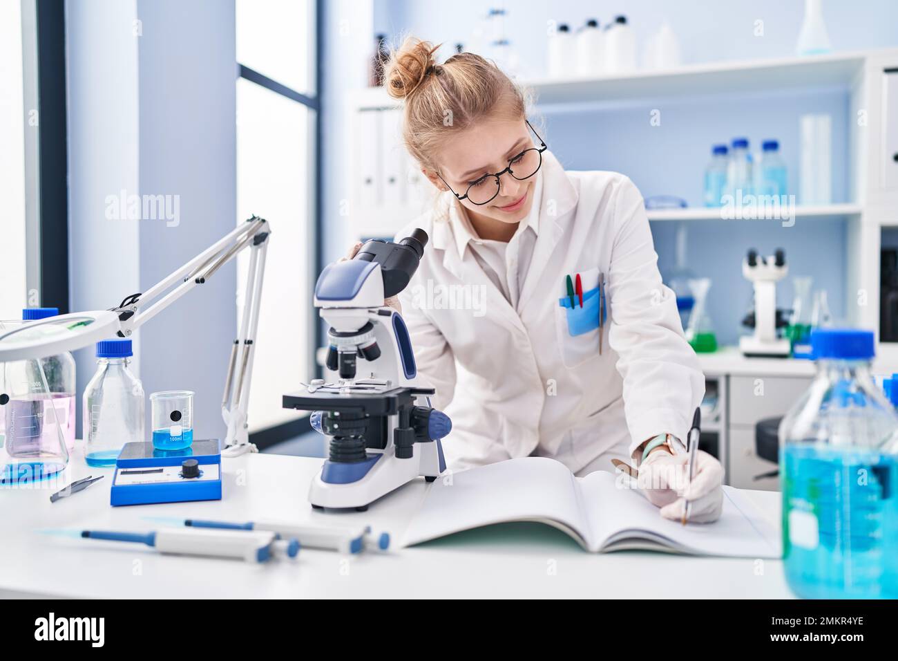 Young blonde woman scientist using microscope writing report at ...