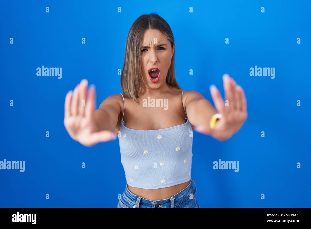 Young woman standing over blue background doing stop gesture with hands ...
