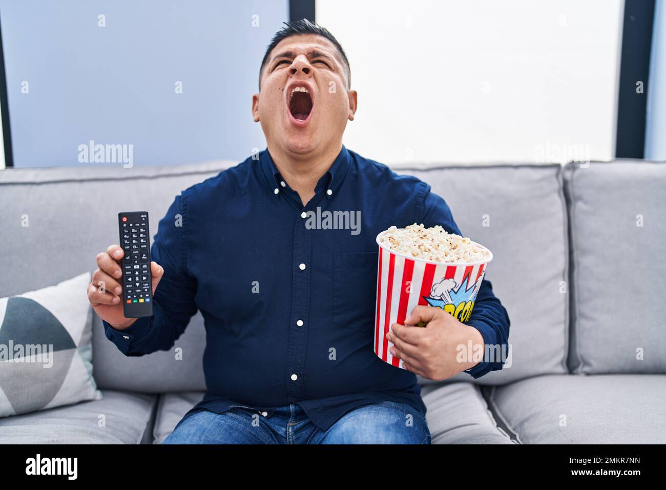 Hispanic young man eating popcorn using tv control angry and mad ...