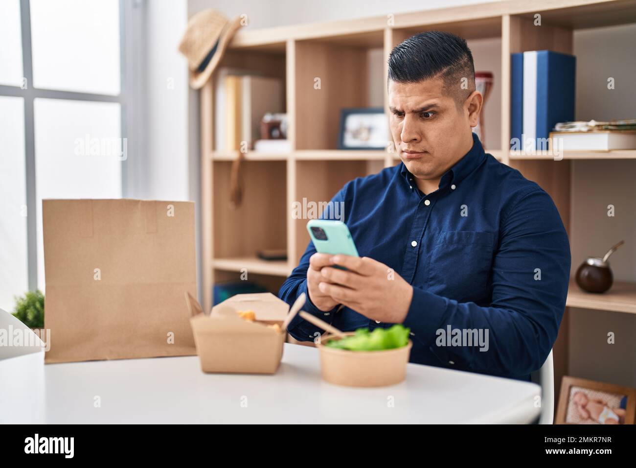 Hispanic young man eating take away food using smartphone skeptic and ...