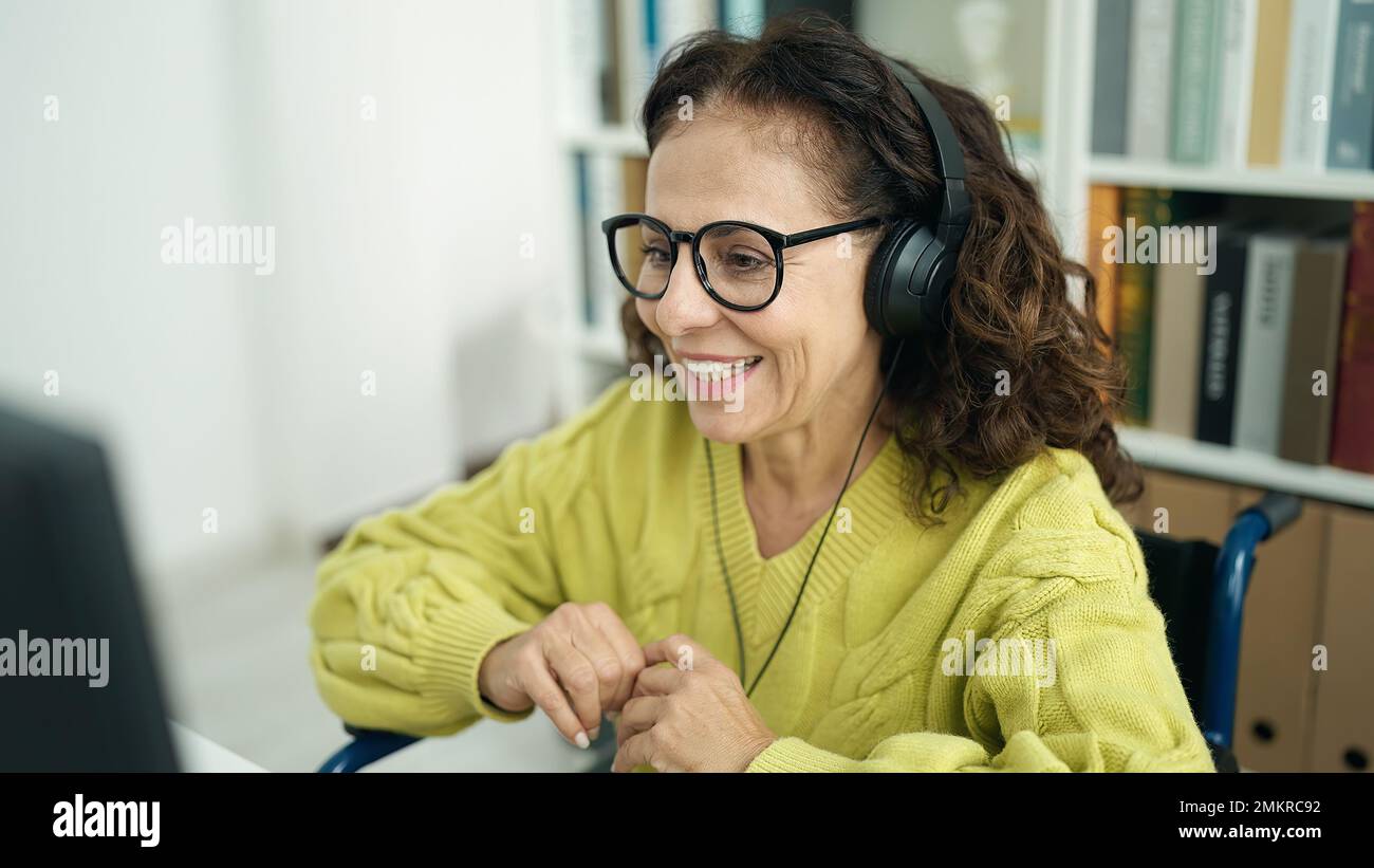 Middle age hispanic woman teacher using computer sitting on wheelchair at library university ...