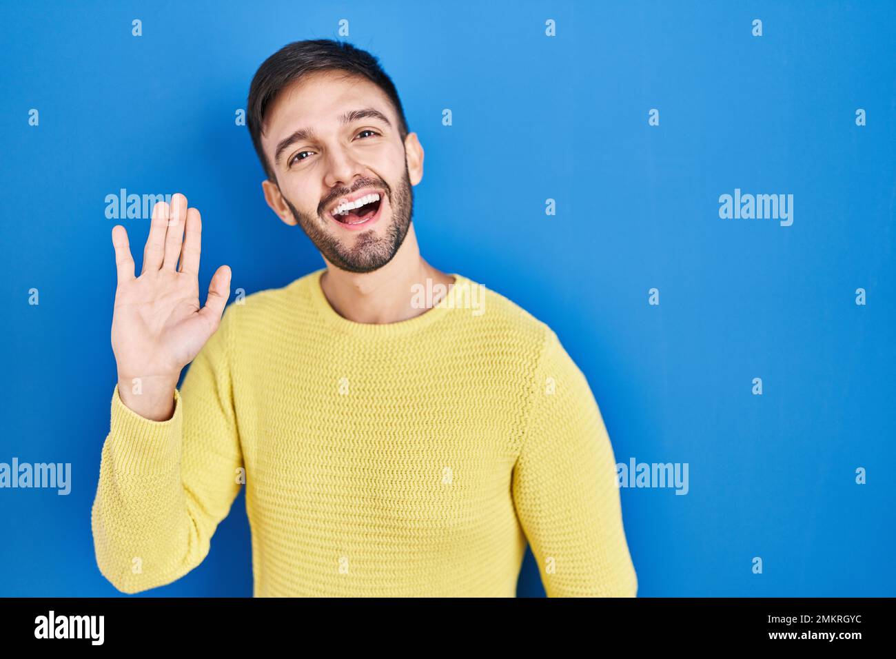 Hispanic man standing over blue background waiving saying hello happy ...