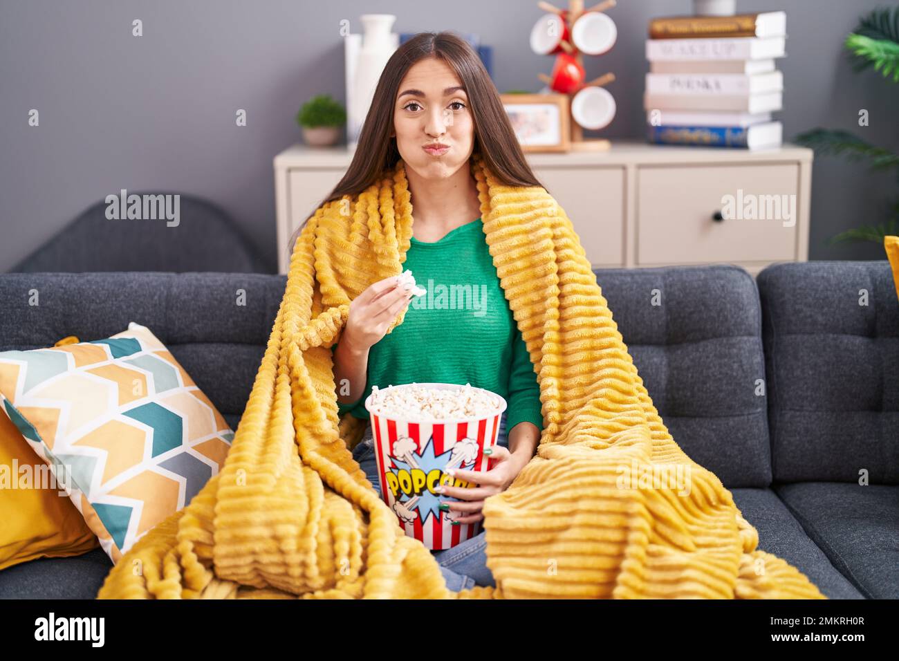 Young hispanic woman eating snack sitting on the sofa at home puffing ...