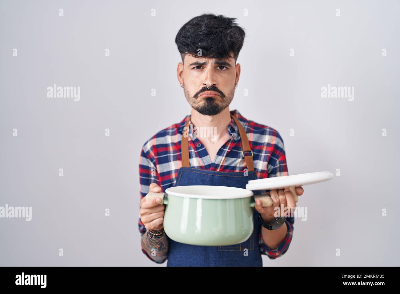 Young hispanic man with beard wearing apron holding cooking pot ...