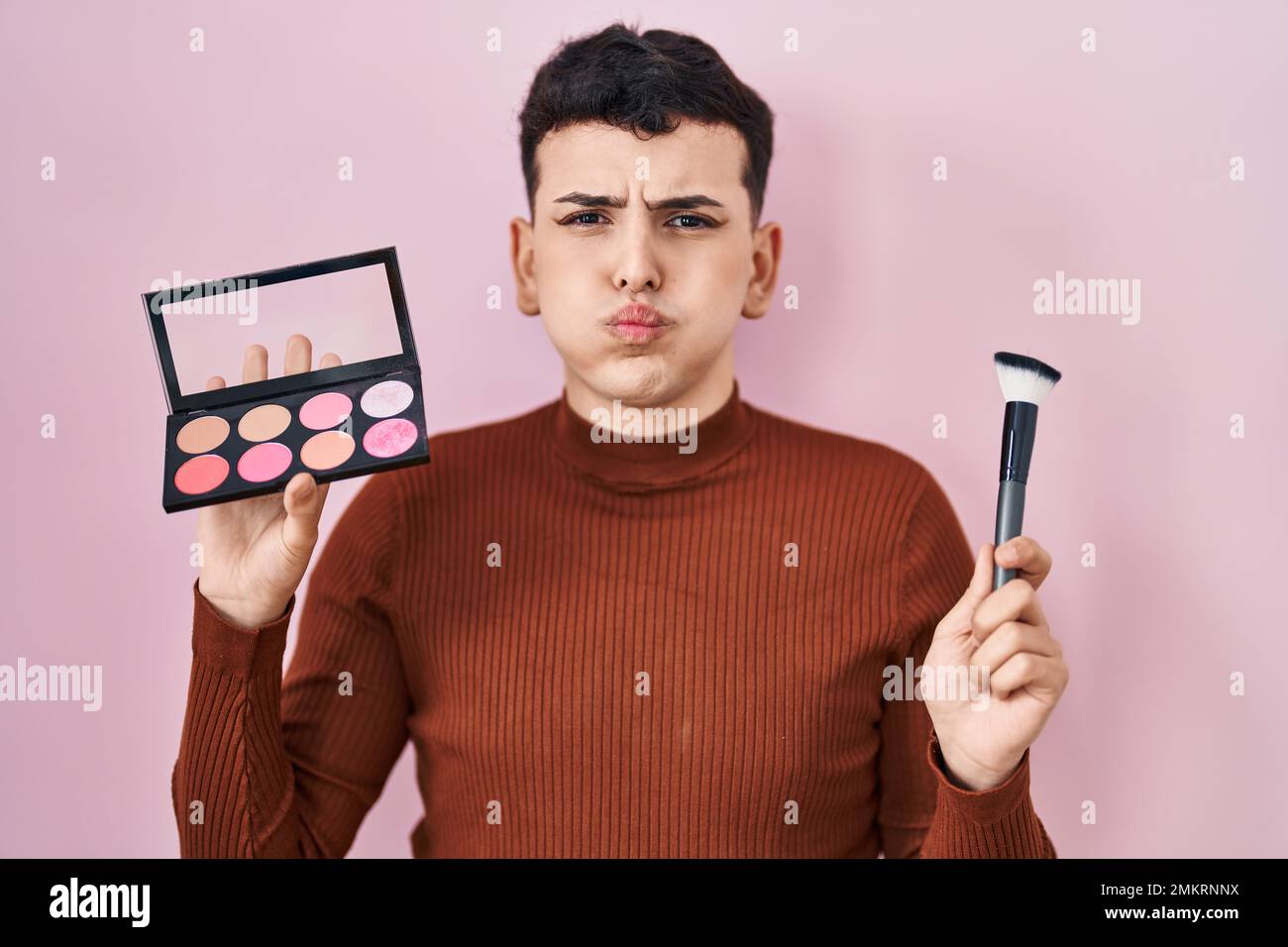 Non binary person holding makeup brush and blush puffing cheeks with ...