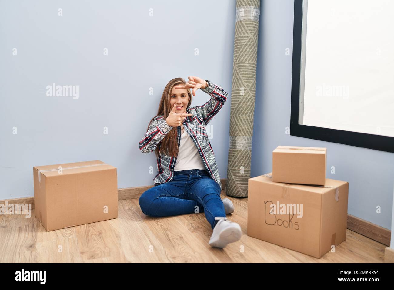 Young blonde woman sitting on the floor moving to a new home smiling ...