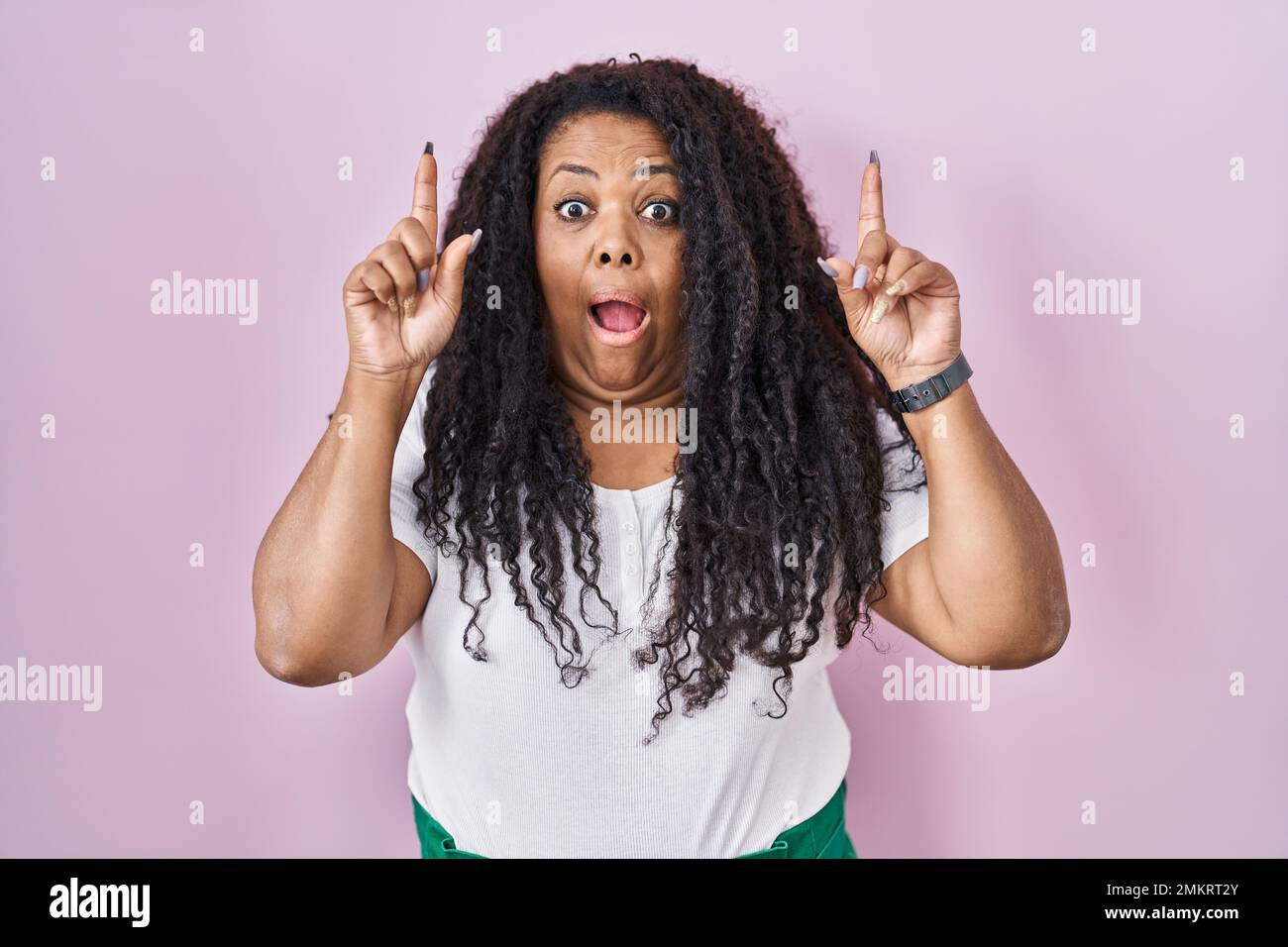 Plus size hispanic woman standing over pink background amazed and ...