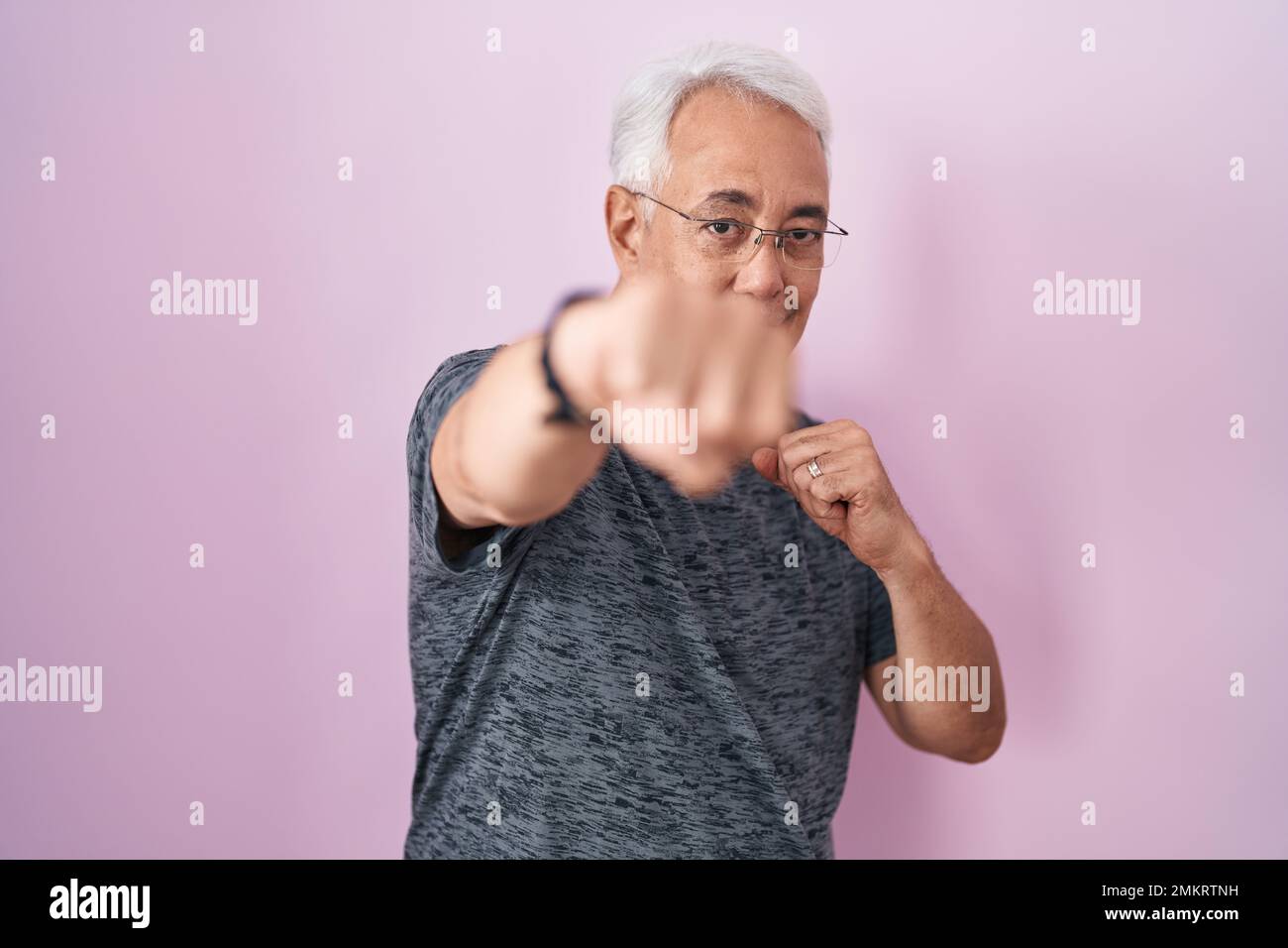 Middle age man with grey hair standing over pink background punching ...