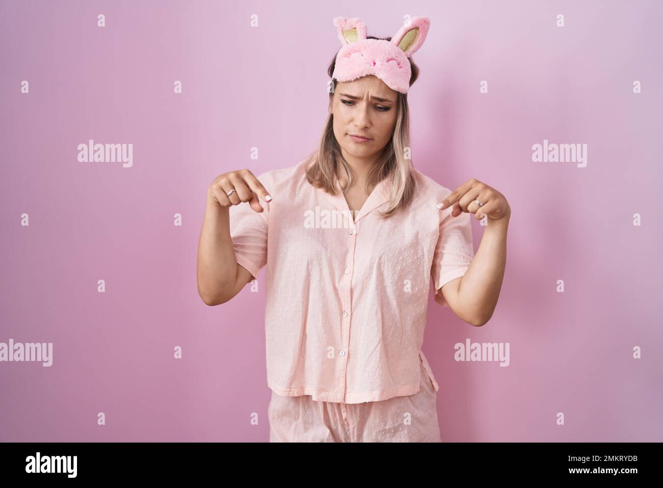 Blonde Caucasian Woman Wearing Sleep Mask And Pajama Pointing Down Looking Sad And Upset
