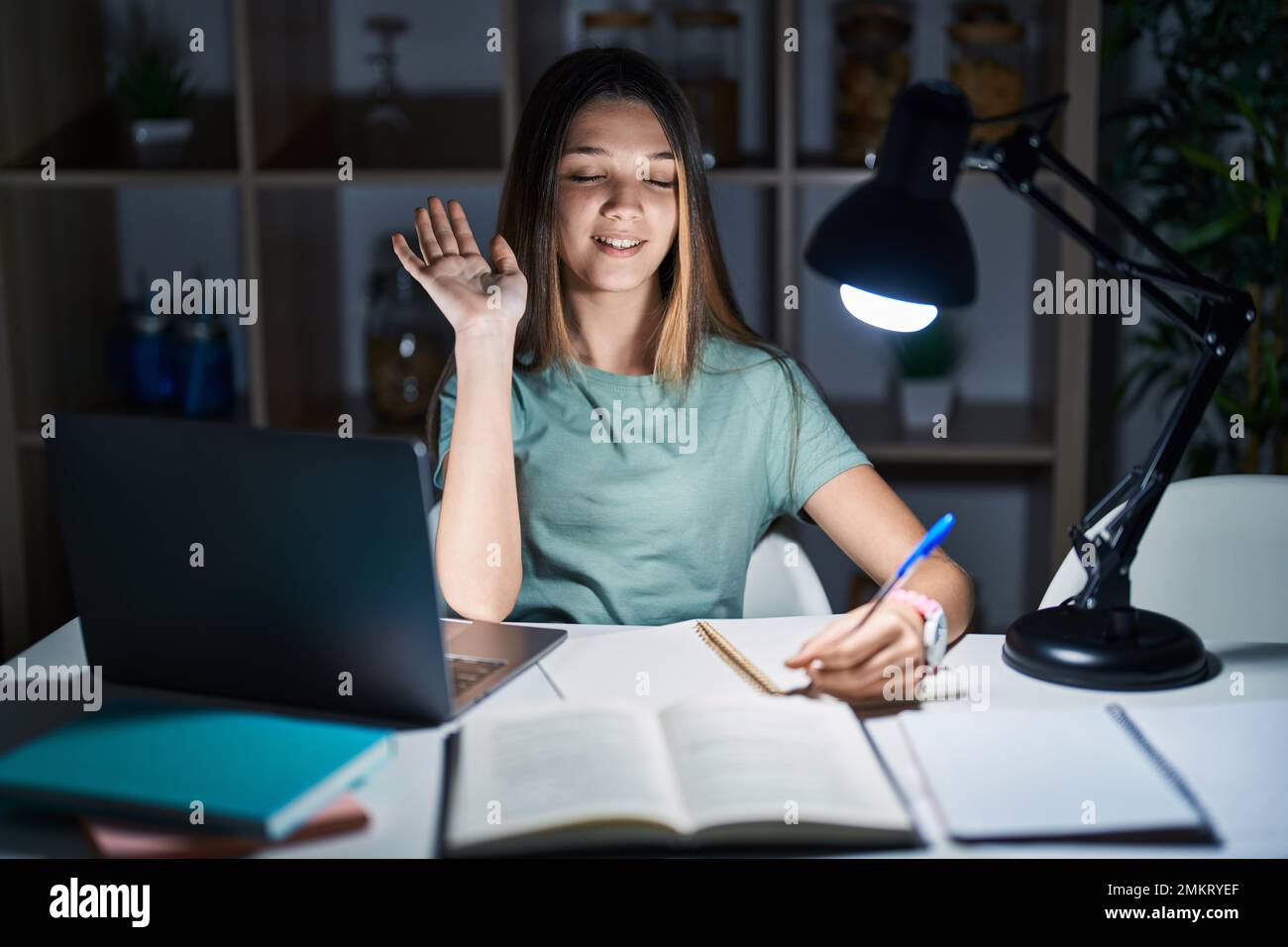 Teenager girl doing homework at home late at night waiving saying hello happy and smiling ...