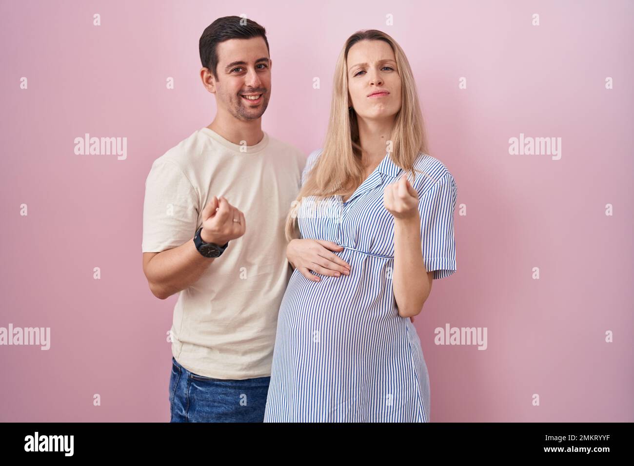 Young couple expecting a baby standing over pink background doing money ...
